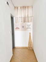 Um corredor com uma porta, uma janela com cortinas leves e três obras de arte emolduradas na parede.