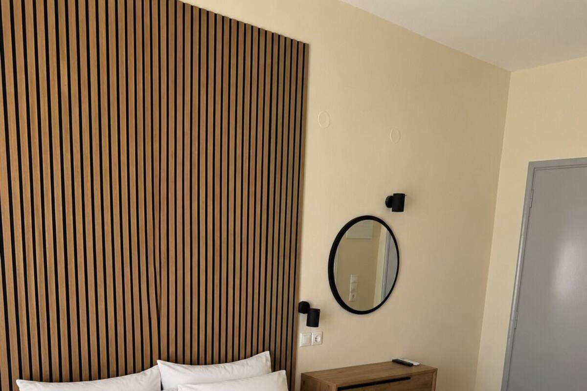 Quarto de hotel com uma cama grande, cabeceira de madeira, mesa de cabeceira com espelho, pequeno armário de madeira e porta cinza.
