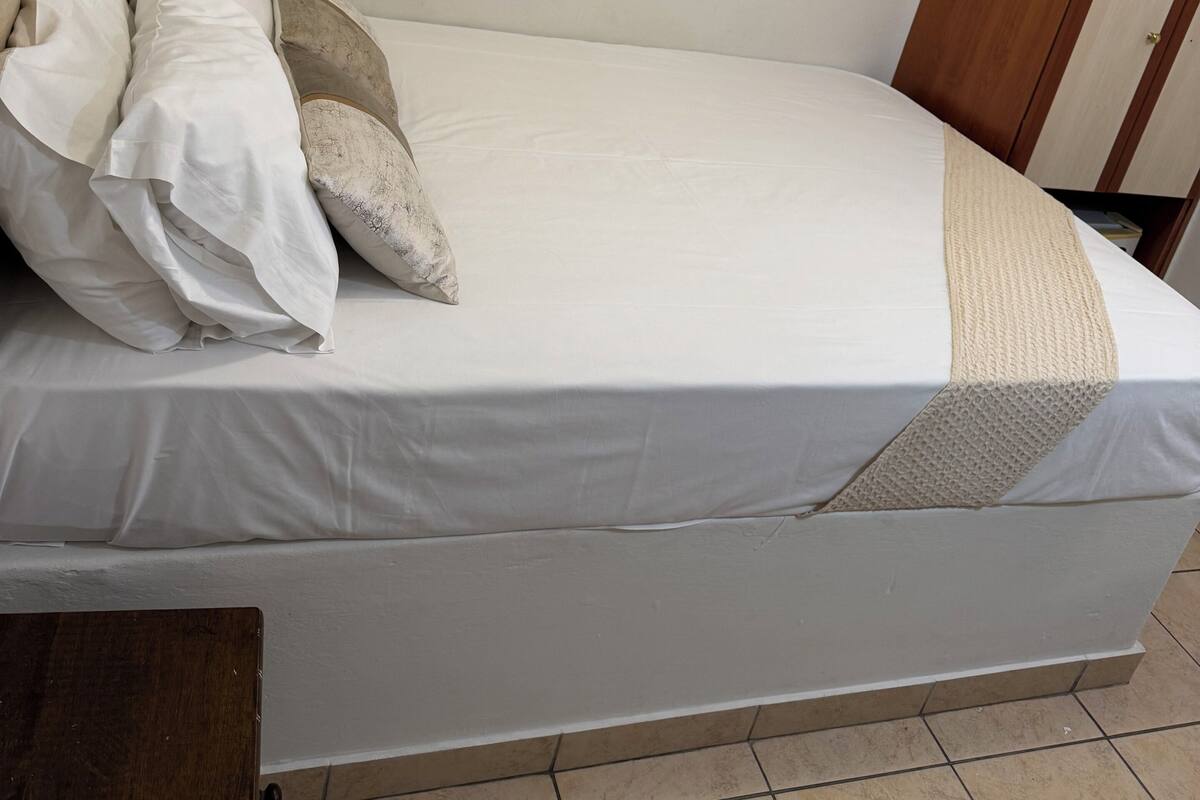 Uma cama de solteiro com colchão branco, manta bege e vários travesseiros.