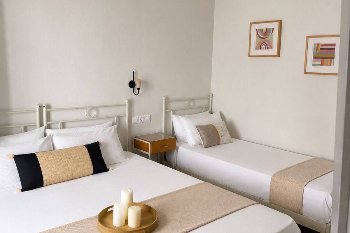 Quarto de hotel com duas camas, uma mesinha de cabeceira, uma luminária na parede e duas obras de arte emolduradas.