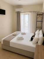 Quarto de hotel com cama, mesa de cabeceira, televisão, ar condicionado, janela com cortinas e vista para edifícios.