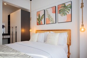 Quarto com cama, roupa de cama branca, cabeceira de vime e três impressões botânicas emolduradas na parede.
