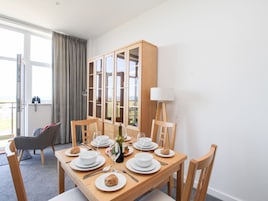 Uma sala de jantar com uma mesa de madeira posta para quatro pessoas, um armário com portas de vidro, um abajur e vista para o exterior.