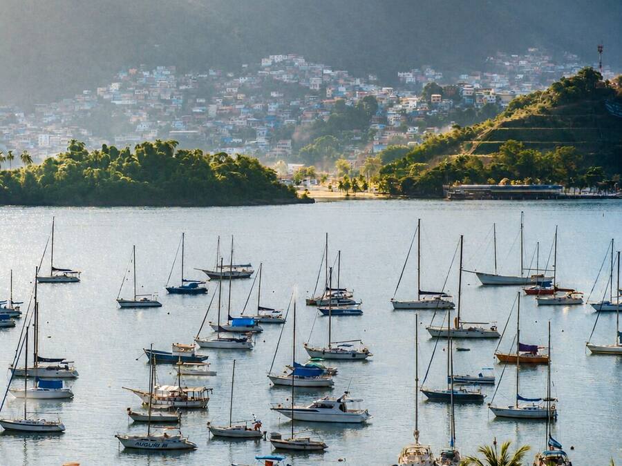 Cena costeira com vários barcos ancorados em águas calmas, com uma povoação densamente povoada ao fundo e palmeiras em primeiro plano.