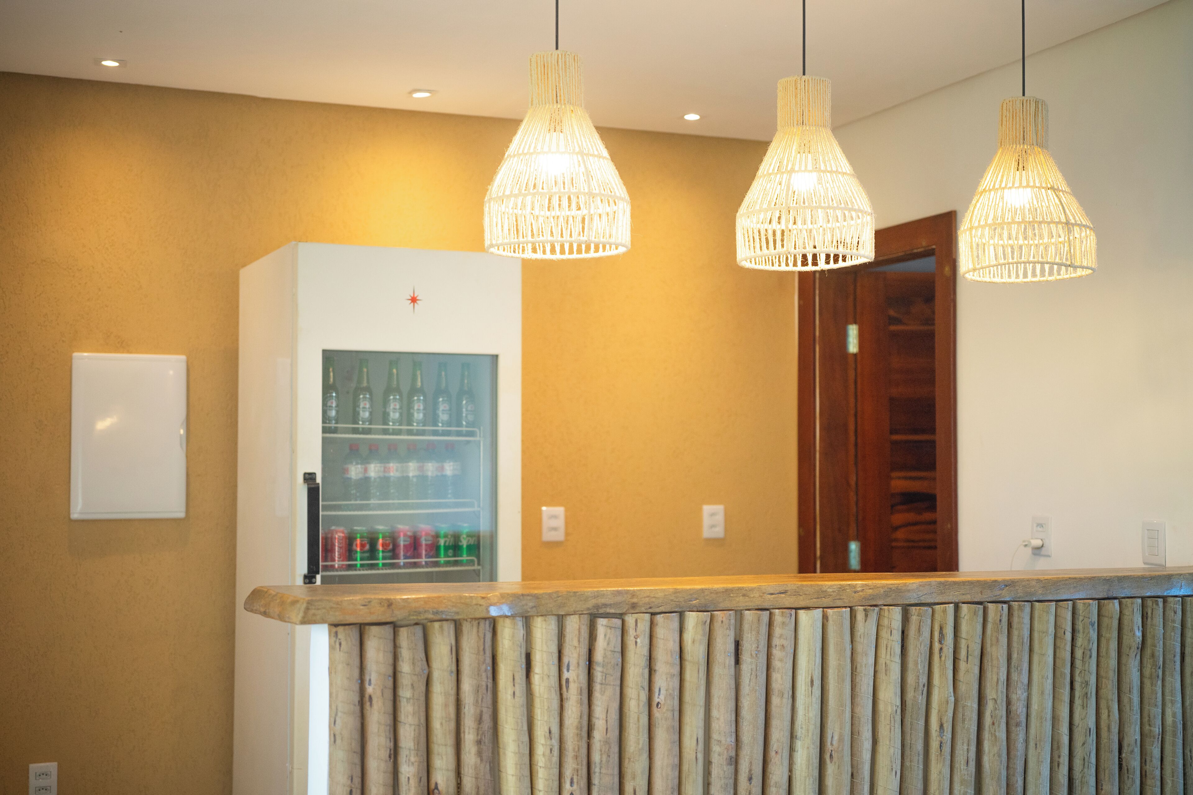 Uma área de recepção com balcão de madeira, uma geladeira abastecida com bebidas e três luminárias pendentes.