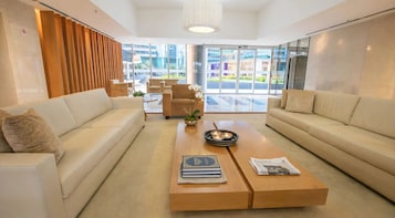 Um hall de hotel moderno com dois sofás, uma mesa de centro com livros e um jornal, e vista da cidade por grandes janelas.