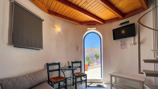Quarto com teto de madeira, televisão fixada na parede, mesa com cadeiras, um sofá e uma porta em arco que dá para o exterior.