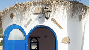 Uma pequena construção com um quarto, porta azul, cama, mesa pequena e janela.