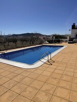 Piscina com deck ladrilhado, rodeada por um jardim ajardinado com árvores e vista para colinas ao longe.
