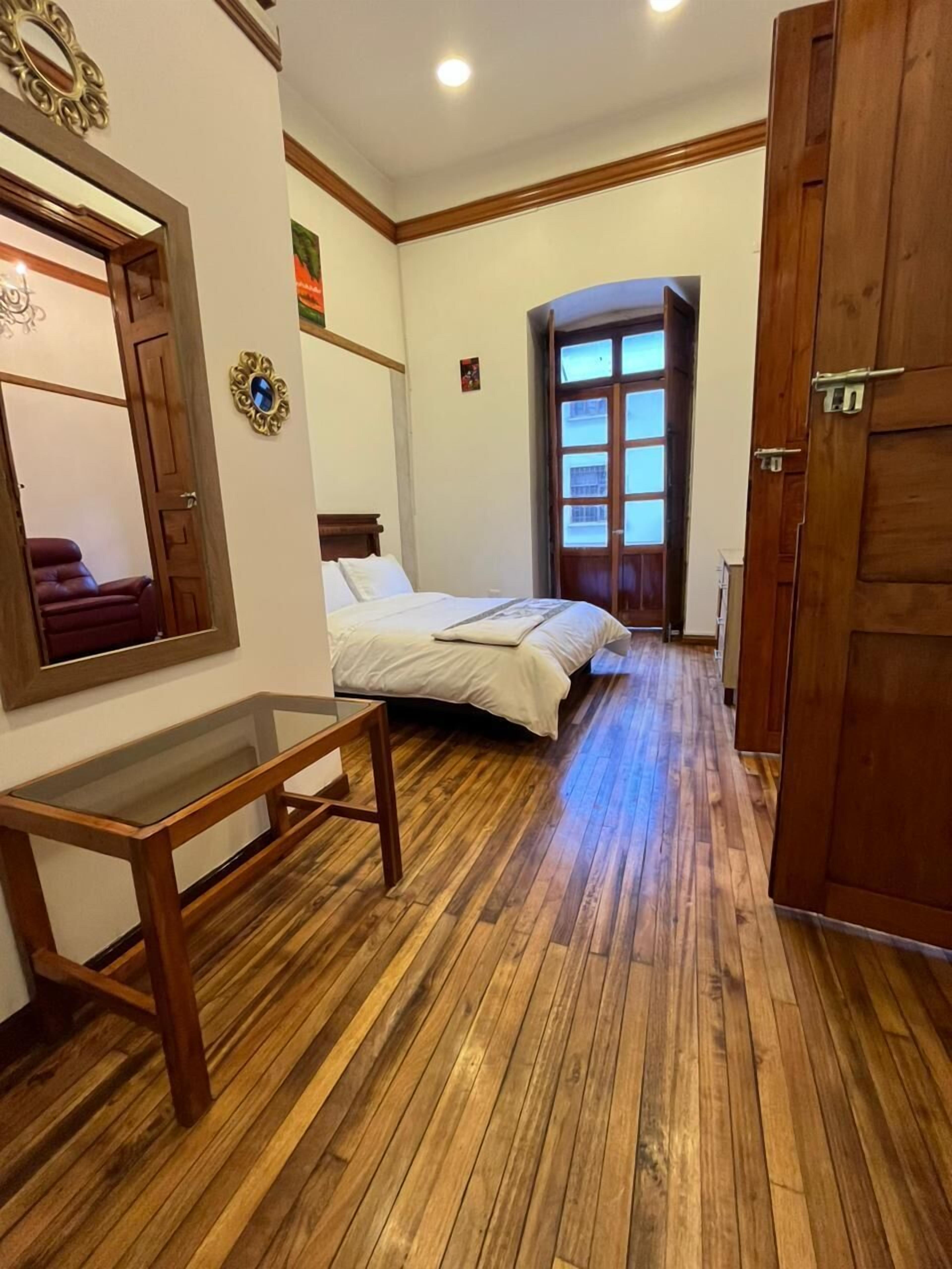 Quarto com piso de madeira, uma cama, uma escrivaninha, uma cadeira e uma porta.