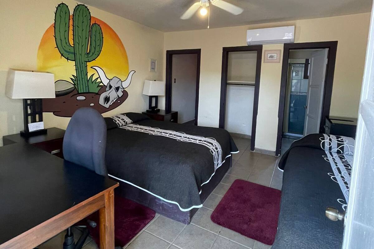 Quarto de hotel com cama, escrivaninha, cadeira, ventilador de teto e um mural de cactos e caveira.