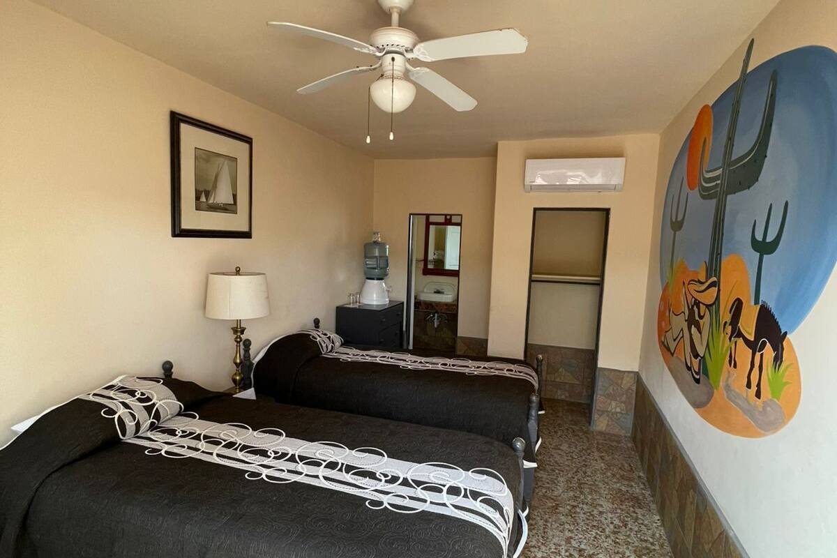 Quarto de hotel com duas camas, um ventilador de teto, uma moldura com imagem, um dispensador de água, um espelho e um mural na parede.