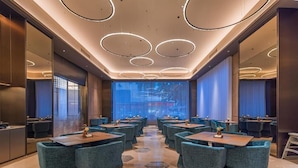 Um hall de hotel moderno com área de estar central, uma tela grande e luminárias circulares no teto.