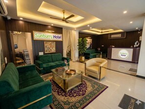 Um hall de hotel com balcão de recepção, sofás verdes, uma mesa de centro e um tapete estampado.