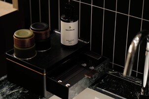 Casa de banho com bancada preta, um frasco de loção corporal Balmain e dois recipientes.