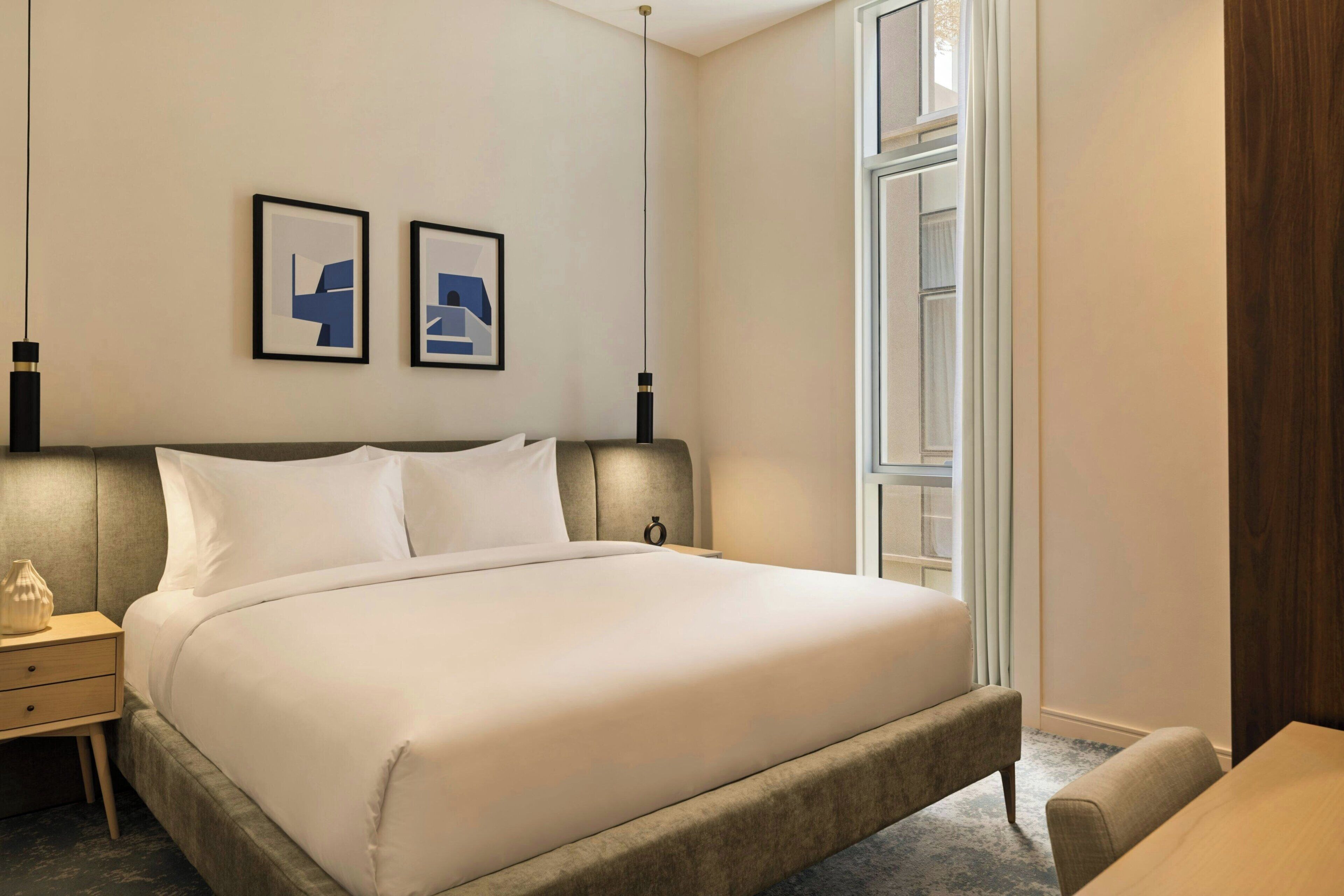Quarto de hotel com uma cama grande, uma mesinha de cabeceira, um criado-mudo e um banco, além de uma janela com cortinas.