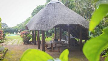 Um pavilhão com telhado de palha, com uma mesa e cadeiras de madeira, rodeado por vegetação exuberante.