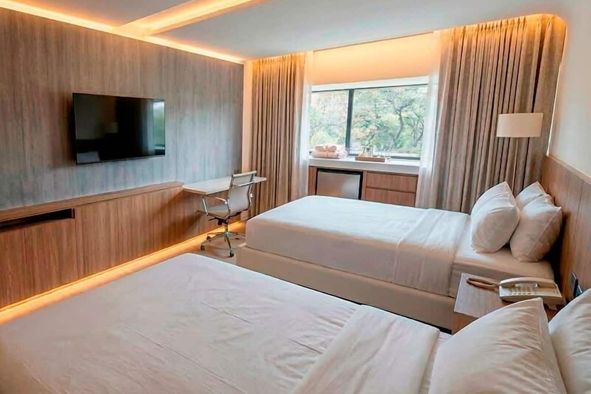 Quarto de hotel com duas camas, uma escrivaninha, uma cadeira, uma televisão e uma janela com cortinas.