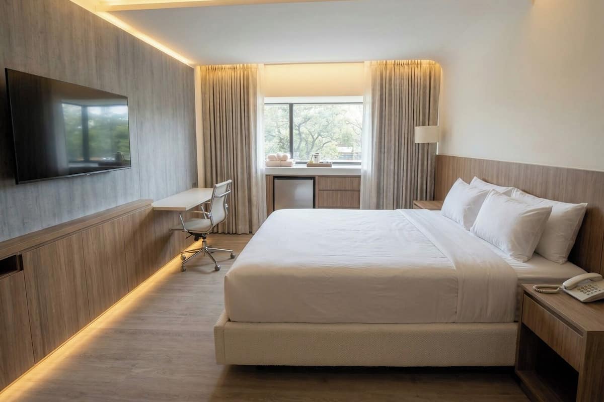 Quarto de hotel com cama, escrivaninha, cadeira, televisão e janela com cortinas.