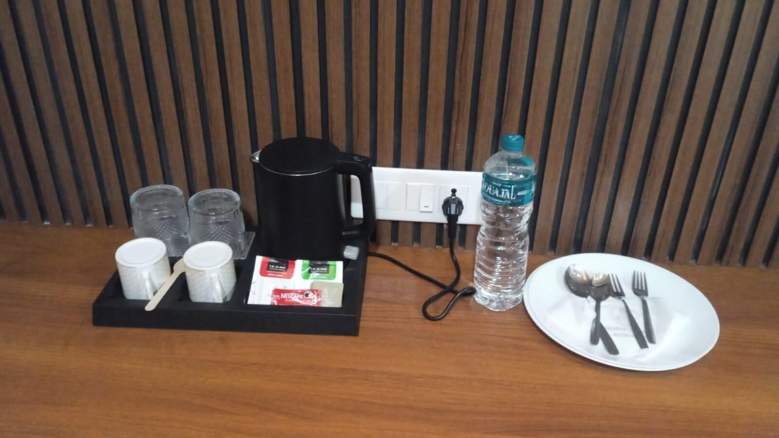 Mesa de quarto de hotel com bandeja preta contendo xícaras, cafeteira, garrafa de água e um prato com talheres.