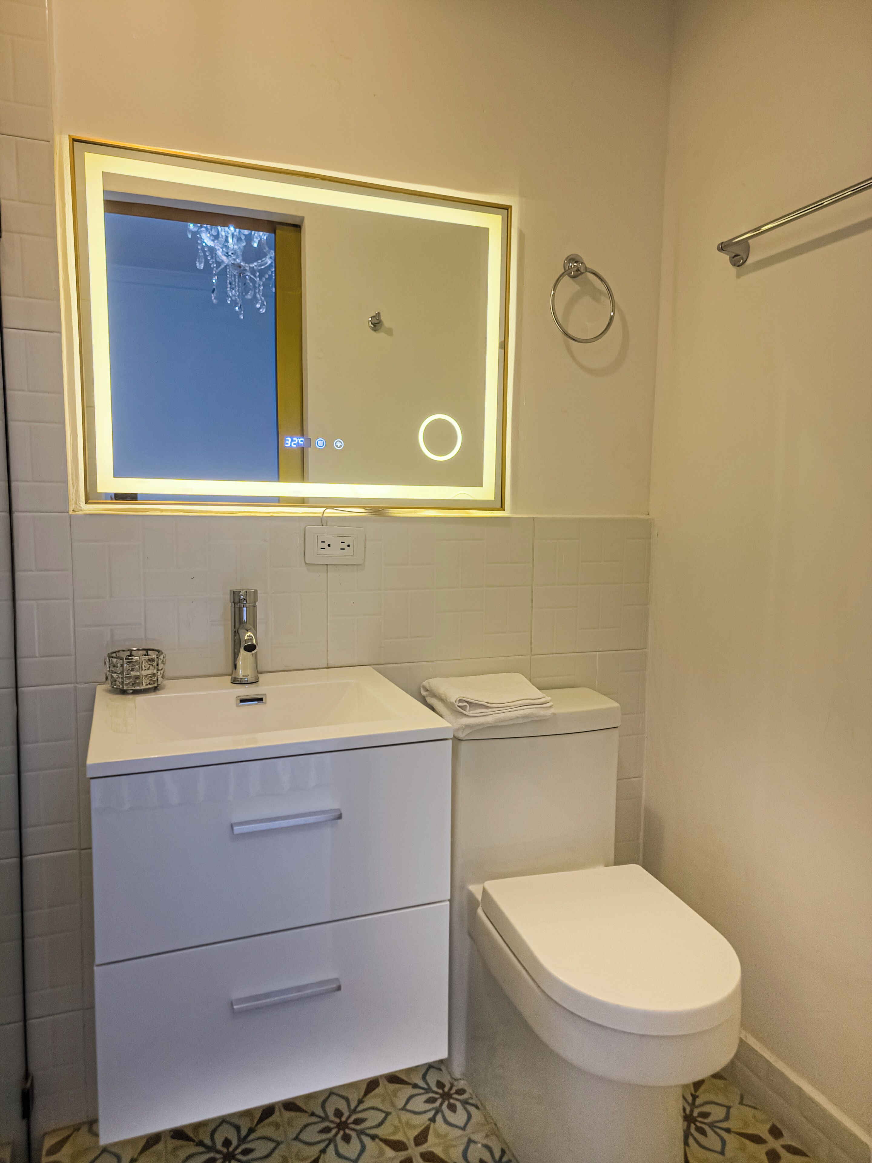 Casa de banho com lavatório branco, sanita, espelho com iluminação nas bordas e porta-toalhas.