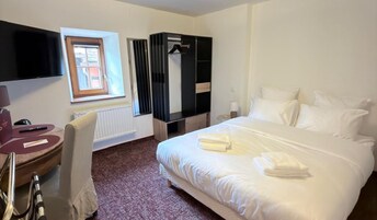Quarto de hotel com cama, escrivaninha, cadeira, televisão, armário e janela.