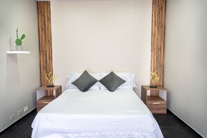 Cama bem arrumada com duas almofadas, uma pequena planta em vaso e duas mesas de cabeceira com objetos decorativos.