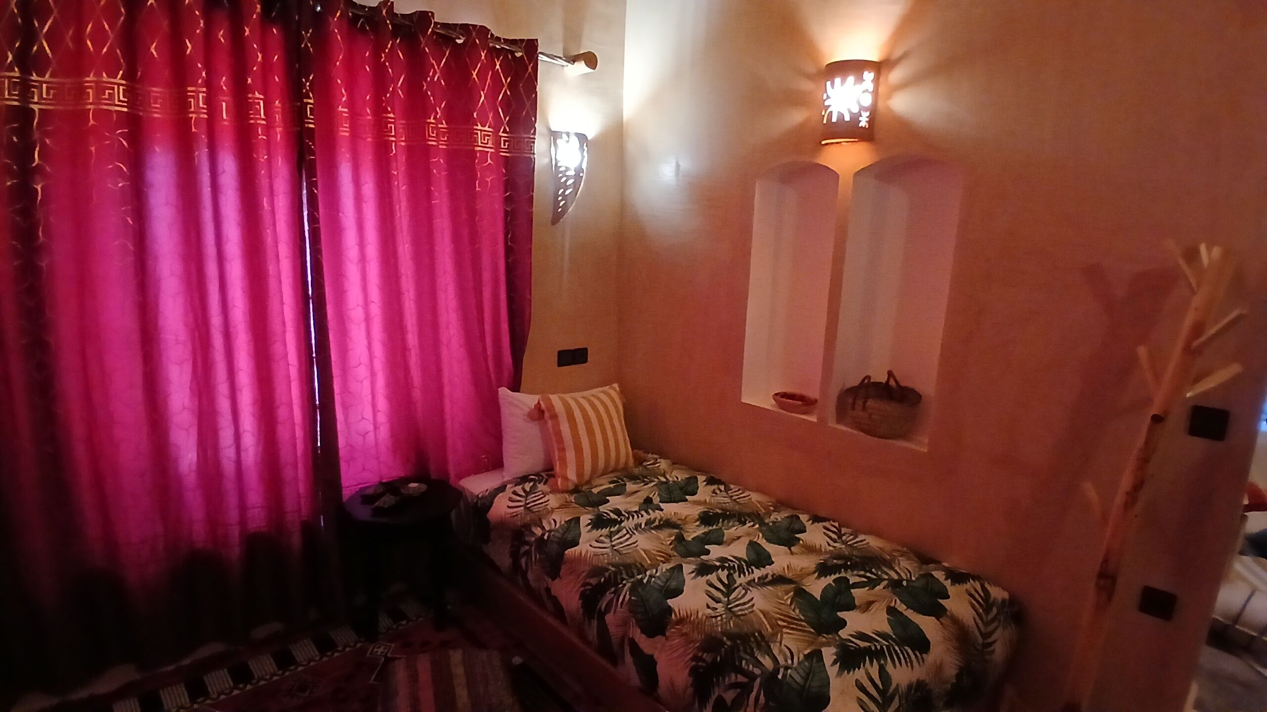 Um quarto com cama, cortinas, uma luminária na parede e uma janela pequena.