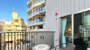Varanda com uma mesa redonda e cadeiras de vime, com vista para uma rua com edifícios.