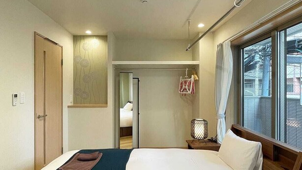 Quarto de hotel com duas camas, uma janela com persianas, uma mesa de cabeceira com abajur e uma porta.