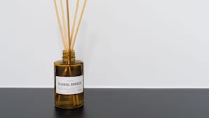 Um frasco de óleo perfumado com palitos de junco, rotulado 'MORHEA FLORAL BREEZE,' sobre uma superfície preta contra um fundo branco.