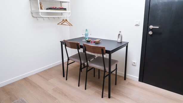 Um quarto pequeno com uma mesa e duas cadeiras, uma estante branca com alguns objetos, e uma porta preta.