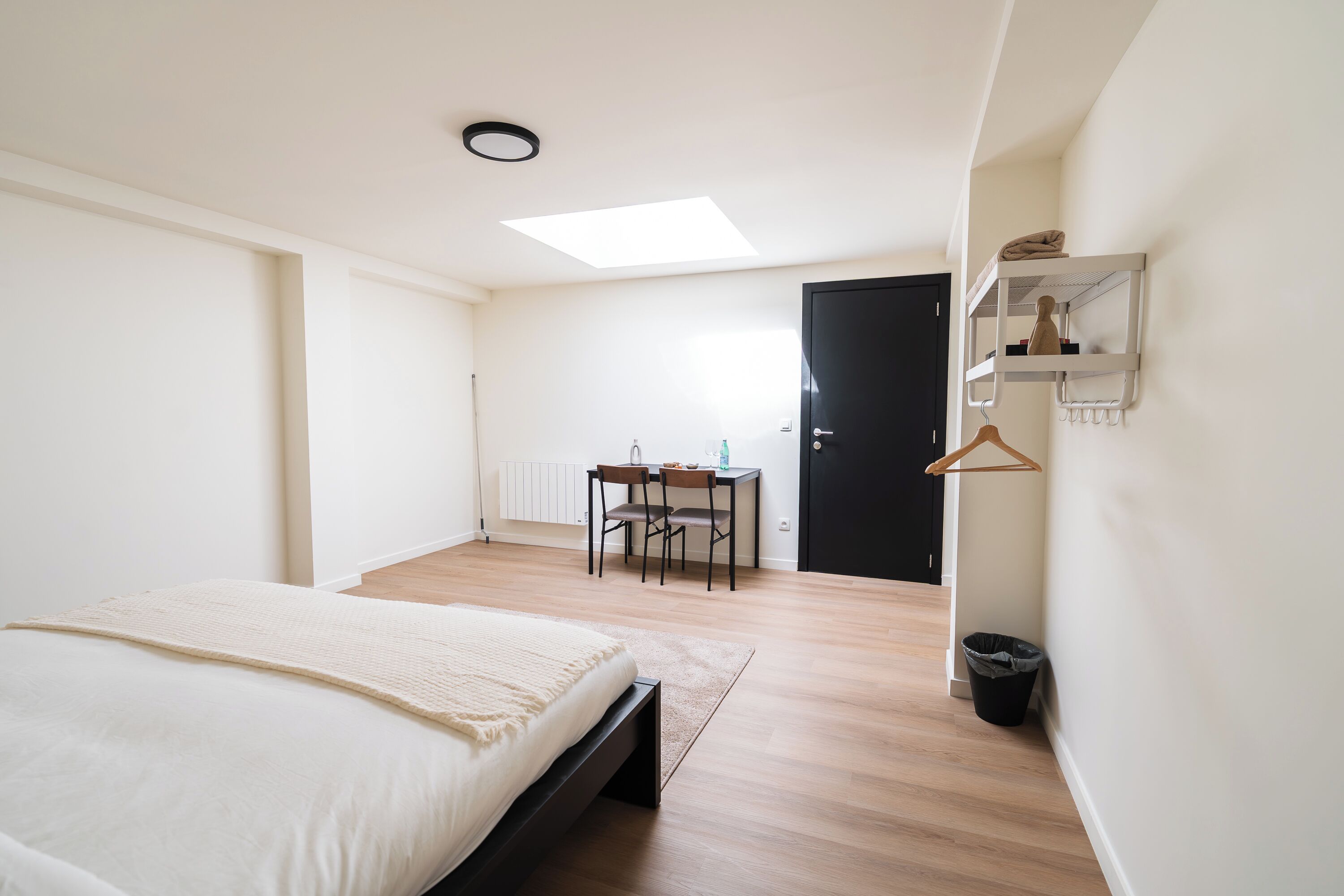 Quarto com cama, escrivaninha, cadeiras, porta preta e radiador.