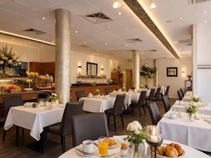 Uma sala de jantar de hotel com mesas montadas para o café da manhã, um buffet com diversas opções de comida e um espaço de bar.