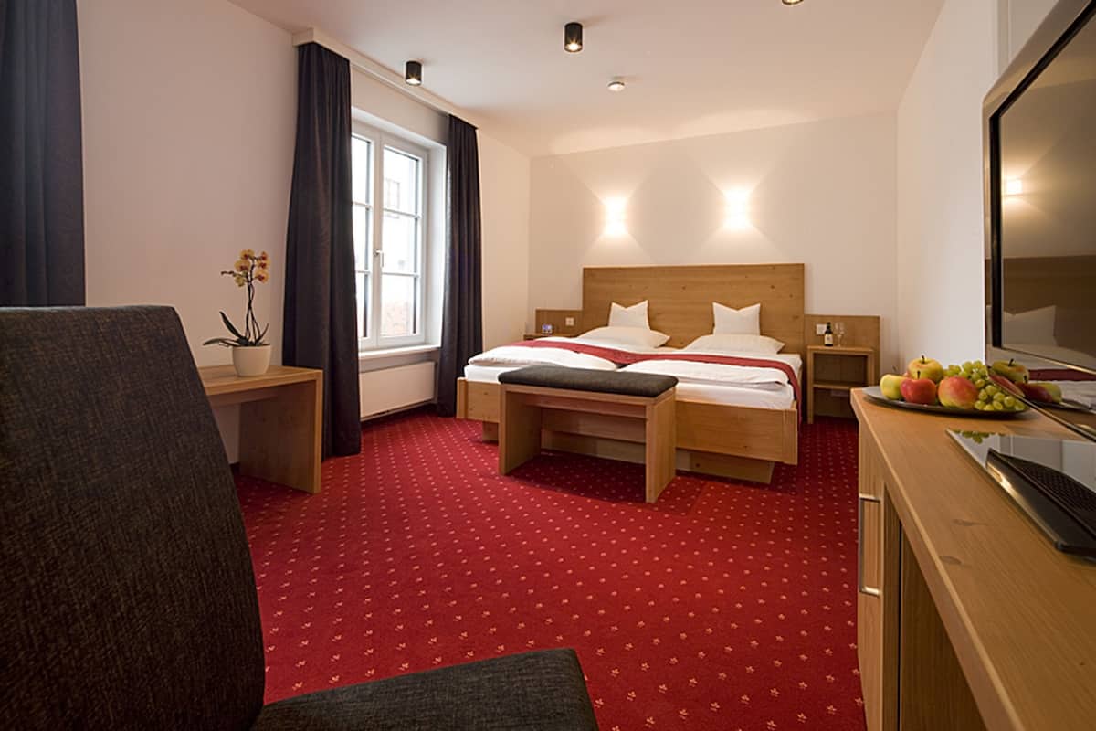 Quarto de hotel com cama, escrivaninha, cadeira, janela com cortinas e uma fruteira sobre a escrivaninha.