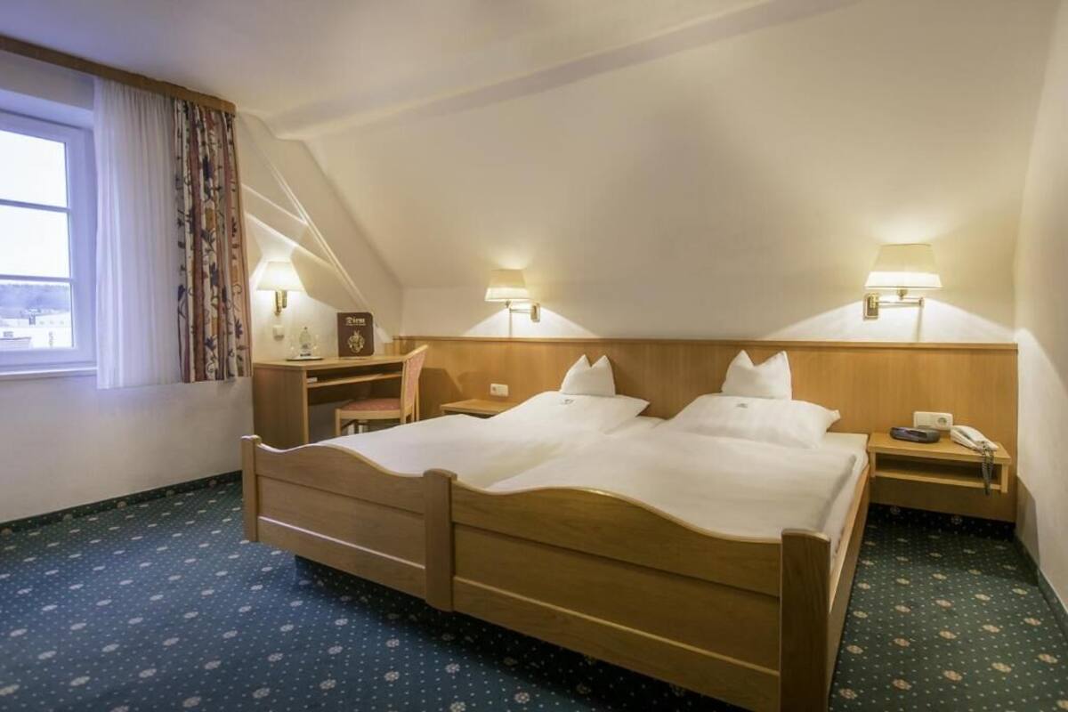 Quarto de hotel com cama de madeira, duas mesas de cabeceira com luminárias, uma escrivaninha e uma janela com cortinas.