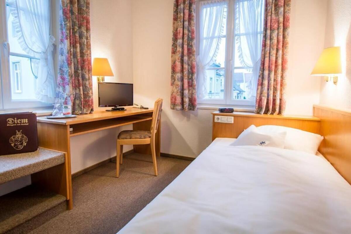 Quarto de hotel com cama, escrivaninha, cadeira, computador e janela com cortinas.
