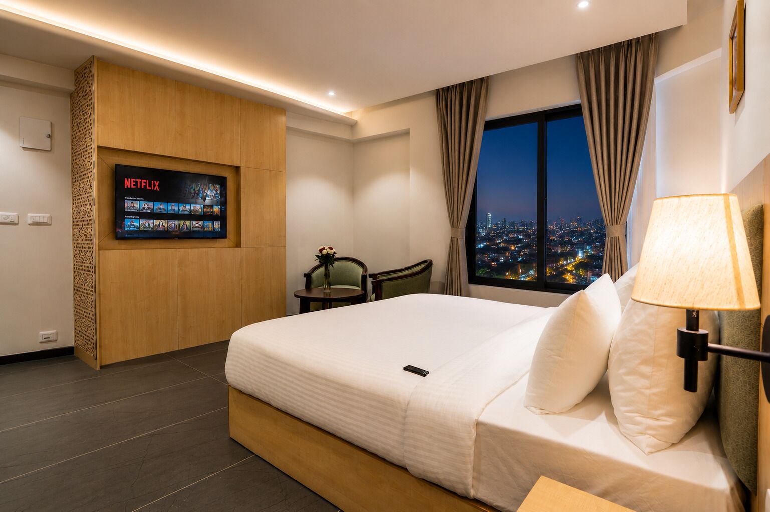 Quarto de hotel com cama, televisão fixada num painel de madeira, vista para a cidade pela janela e mesa de cabeceira com luminária.