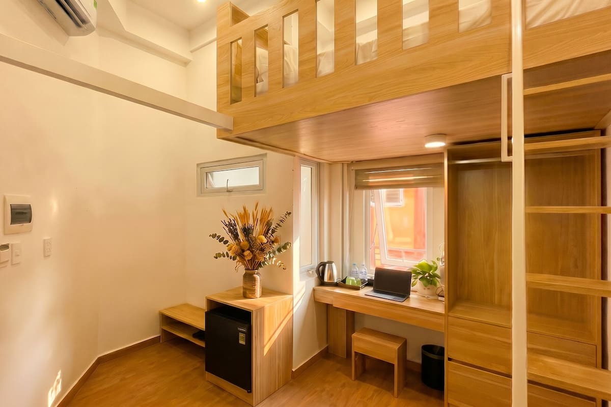 Quarto compacto com beliche, escrivaninha com um laptop, mini geladeira, janela com vista e uma planta.