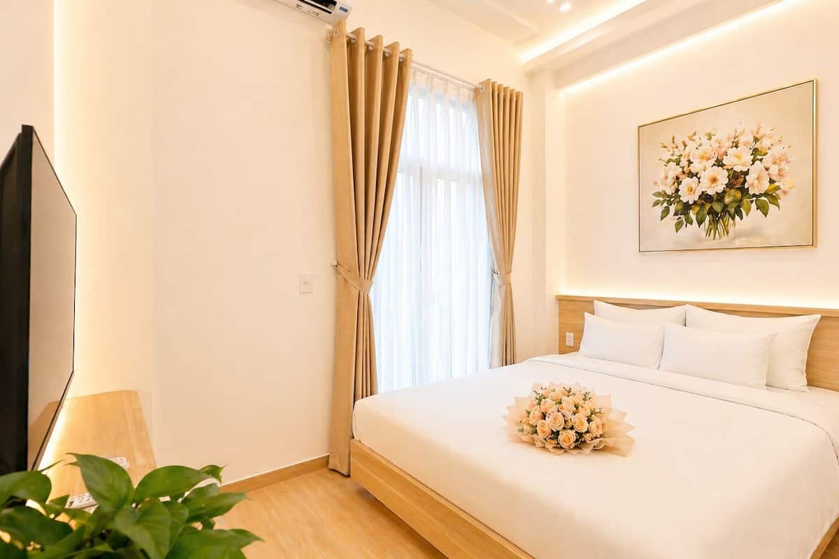Quarto de hotel com uma cama, televisão, janela com cortinas, quadro na parede e planta na mesinha de cabeceira.