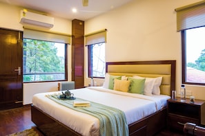 Quarto de hotel com uma cama grande, cabeceira de madeira, mesa de cabeceira, persianas e vista para árvores.