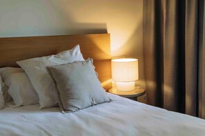 Uma cama bem arrumada com lençóis e almofadas brancos, um abajur e uma cabeceira de madeira.