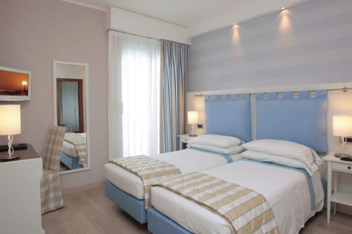 Quarto de hotel com duas camas, uma escrivaninha, uma cadeira, um espelho e uma janela com cortinas.