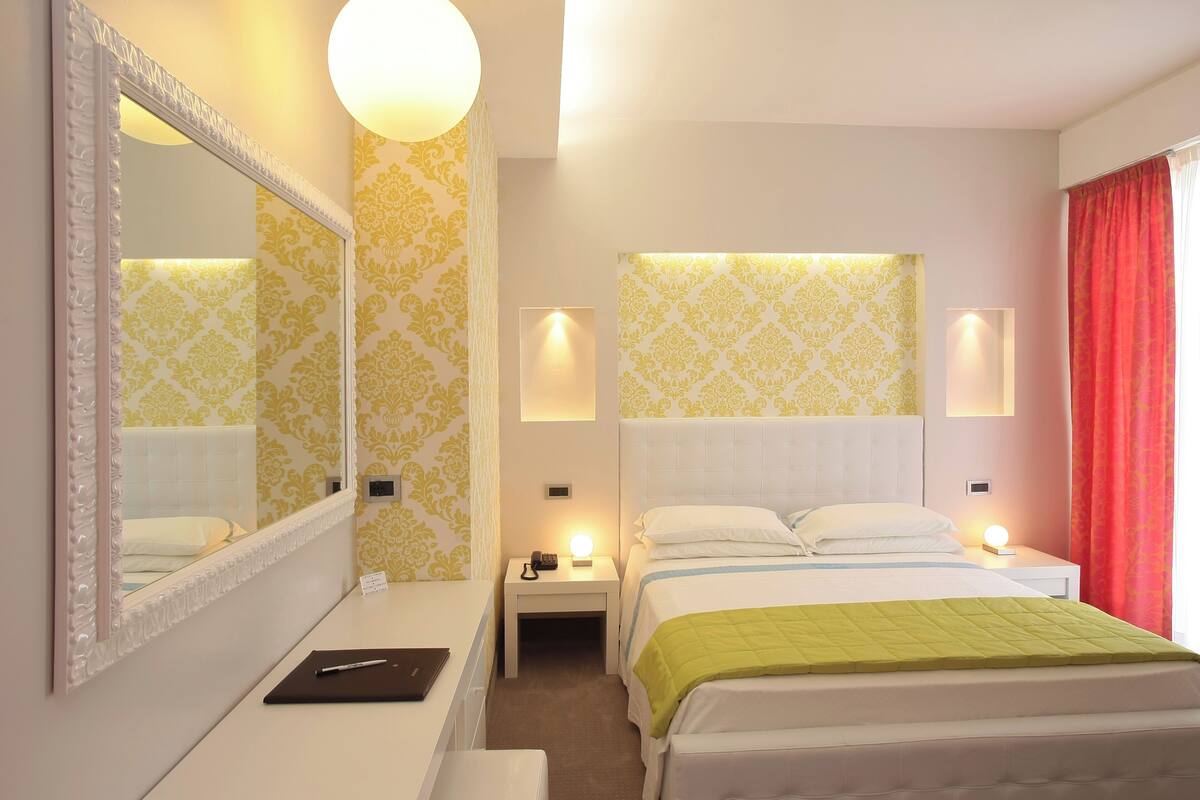 Quarto de hotel com cama, mesas de cabeceira, espelho e escrivaninha com cadeira.