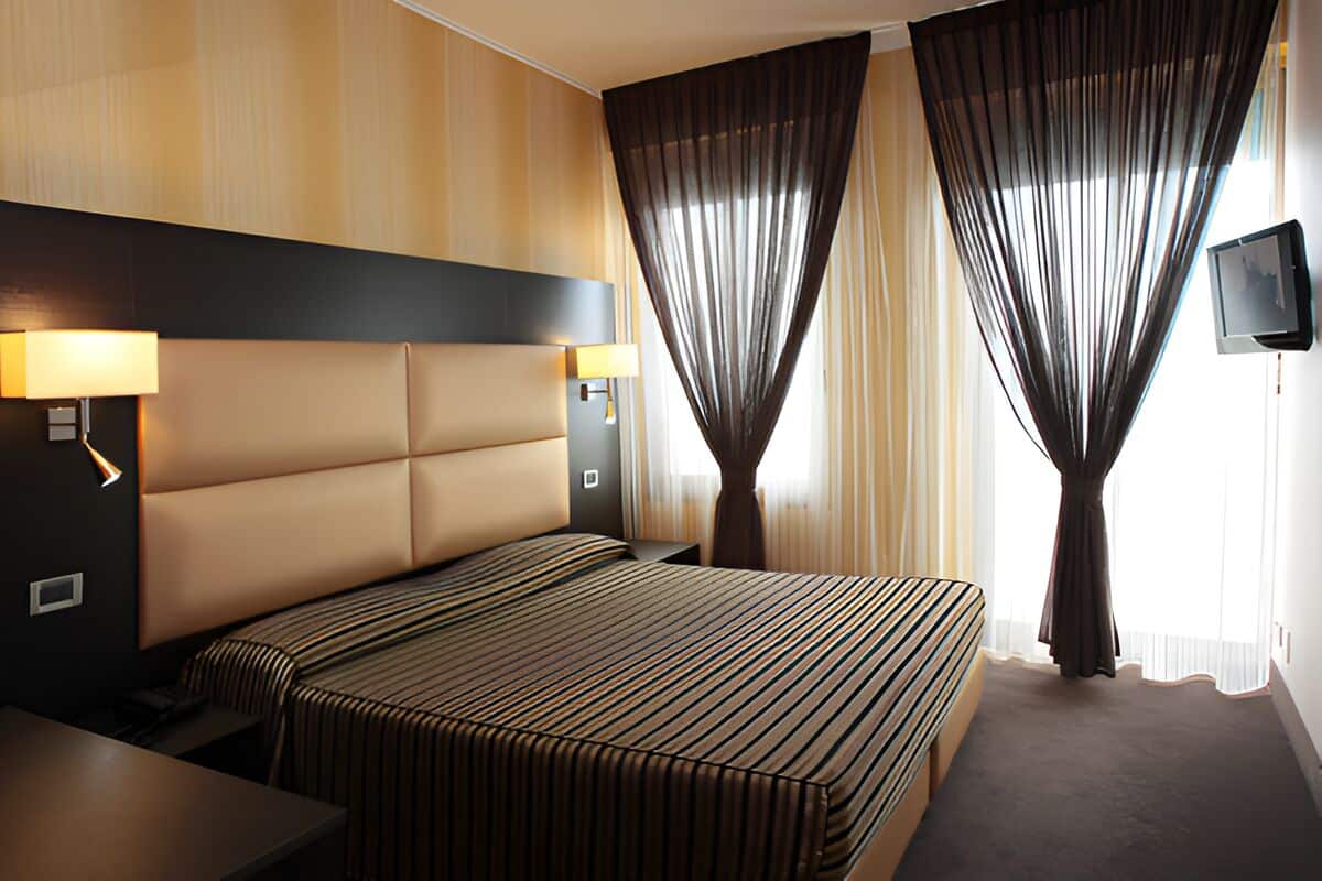 Quarto de hotel com uma cama grande, mesa de cabeceira, televisão e cortinas.