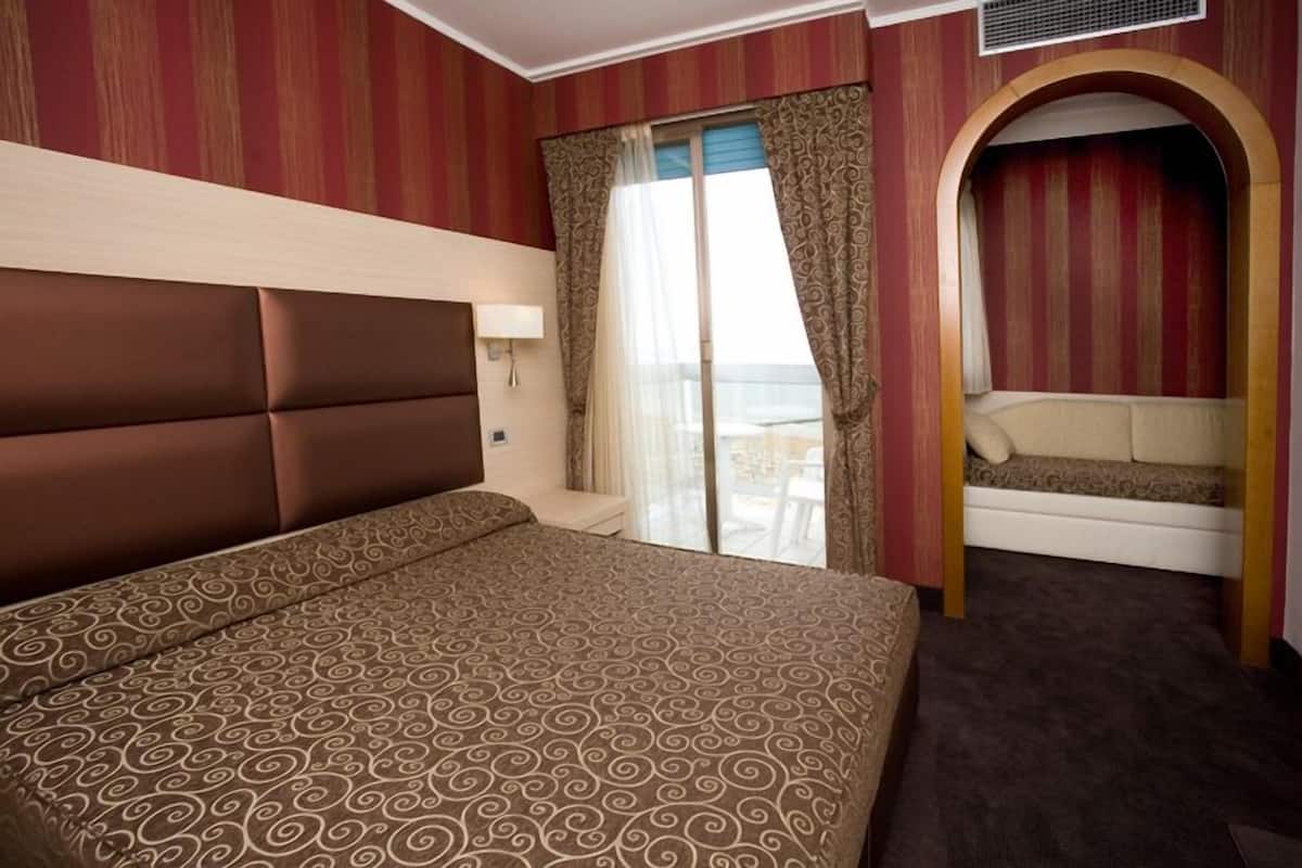 Quarto de hotel com cama grande, sofá, janela com cortinas e vista para o exterior.