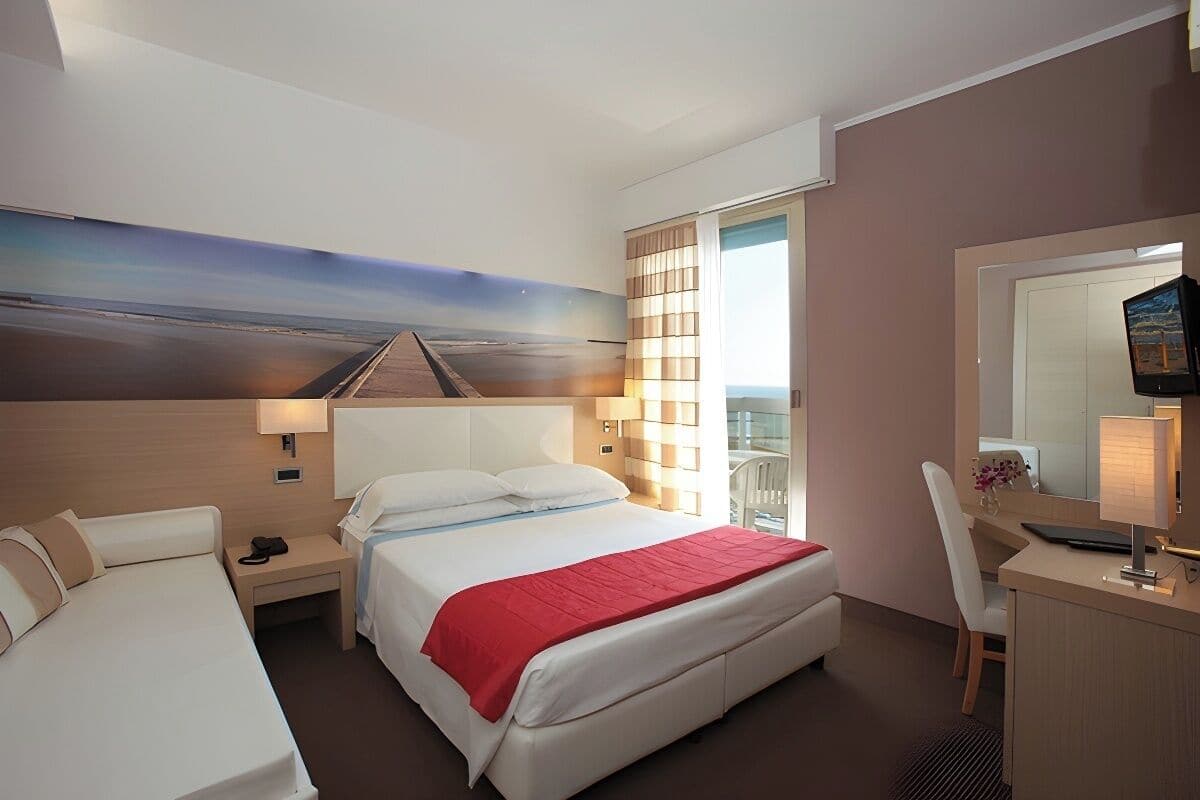 Quarto de hotel com cama, escrivaninha, cadeira, televisão e um quadro de uma paisagem praiana.