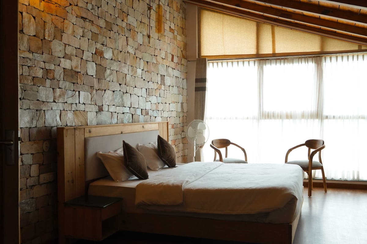 Quarto com parede de pedra, cama de madeira, duas cadeiras e uma janela grande com cortinas.