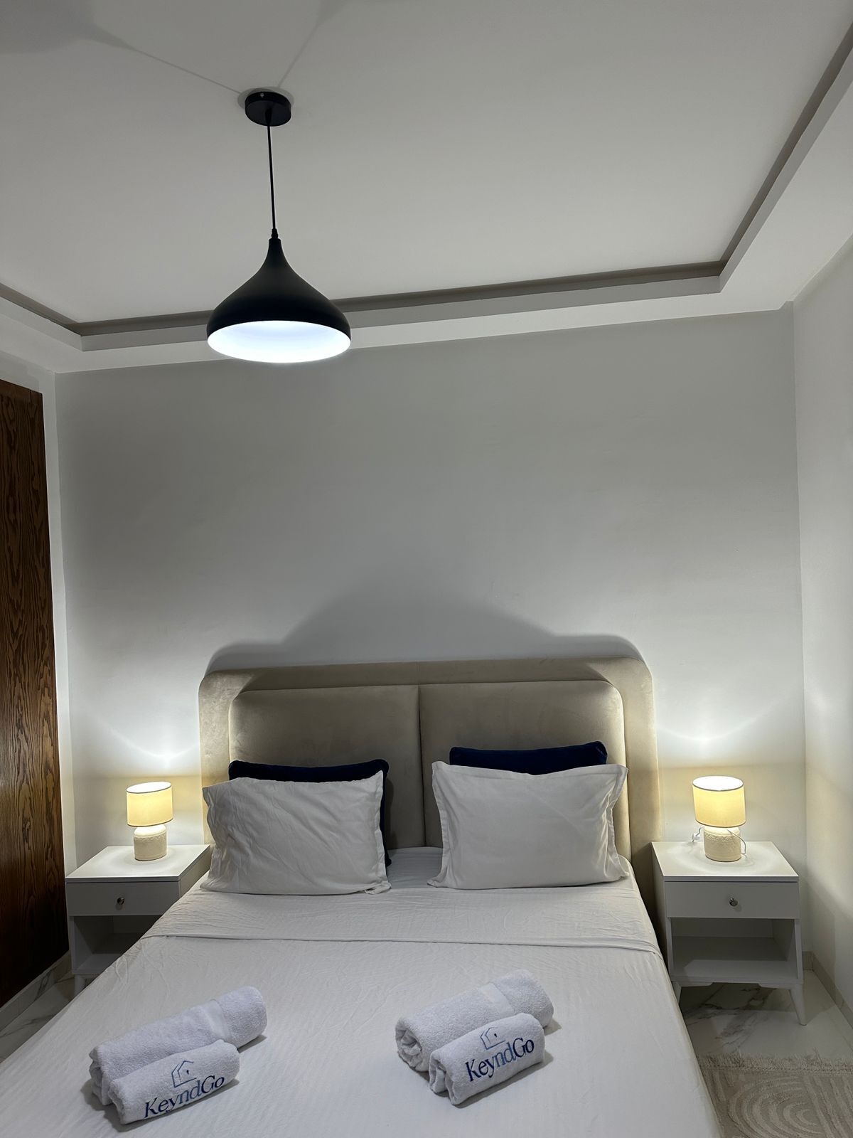 Quarto de hotel com uma cama, duas mesas de cabeceira com luminárias, uma luz no teto e duas toalhas com a marca 'KeyndGo'.