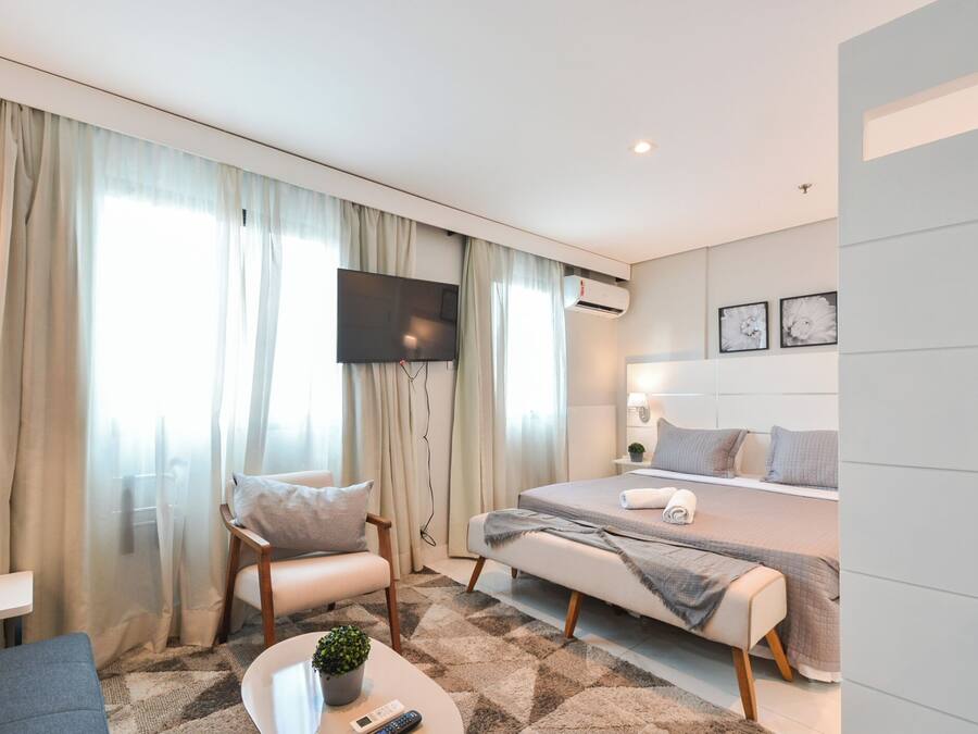 Quarto de hotel moderno com cama, televisão, sofá, mesa de centro com uma planta e uma janela grande com cortinas.